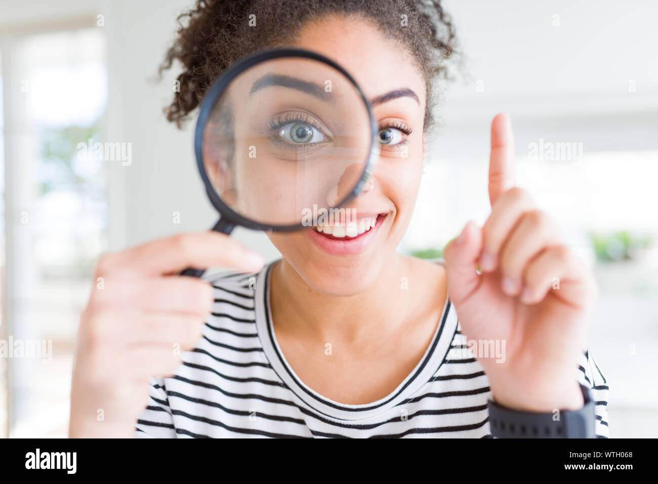 Young african american woman looking through magnifying glass surprised ...