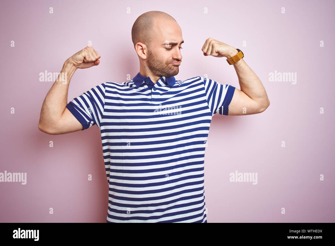 Young bald man with beard wearing casual striped blue t-shirt over pink ...