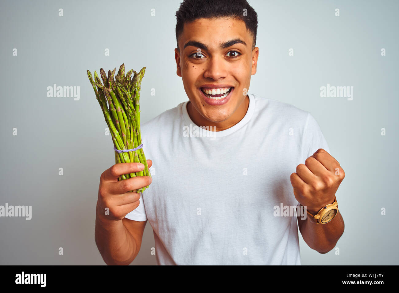 Young brazilian man eating asparagus standing over isolated grey ...