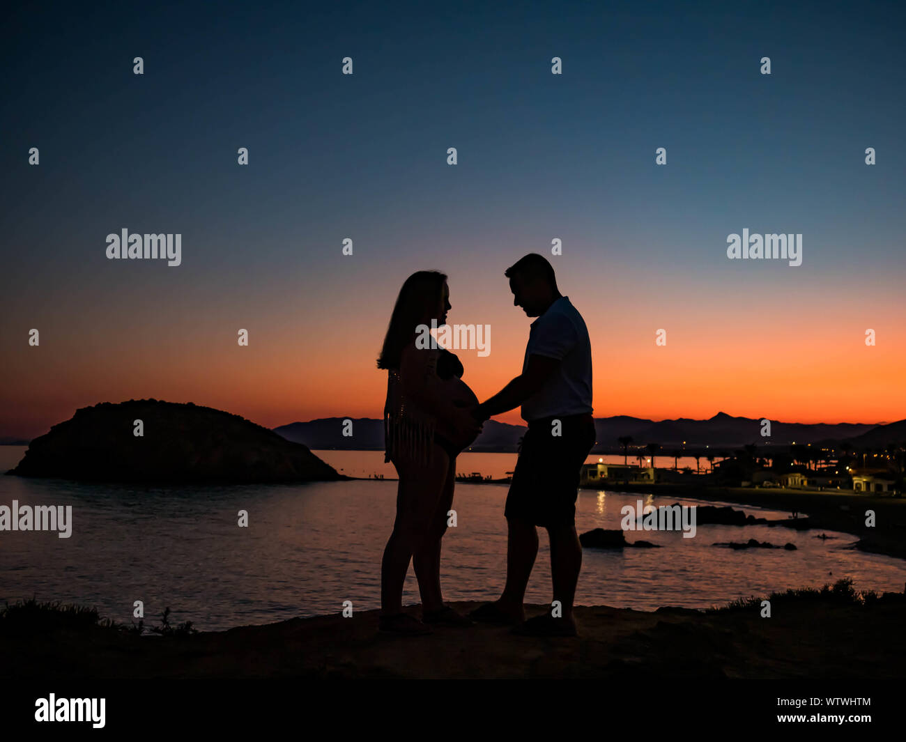 Silhouette of a pregnant couple on a cliff with sunset and city lights ...