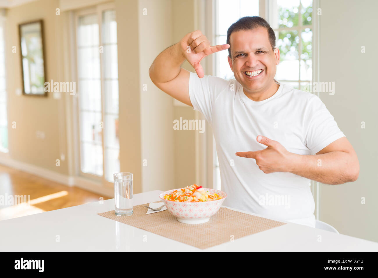 Middle age man eating rice at home smiling making frame with hands and ...