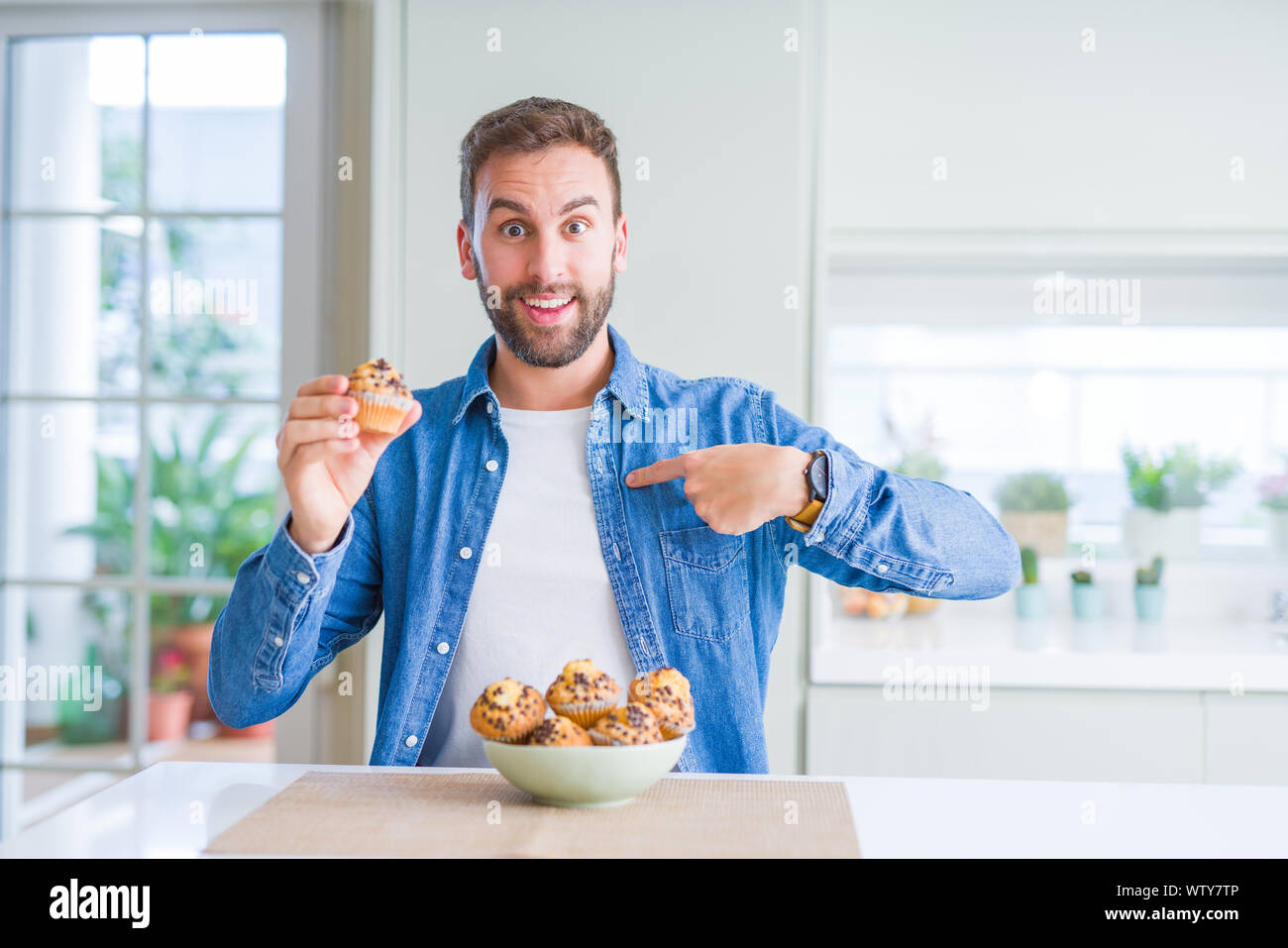 Handsome man eating chocolate chips muffin with surprise face pointing ...