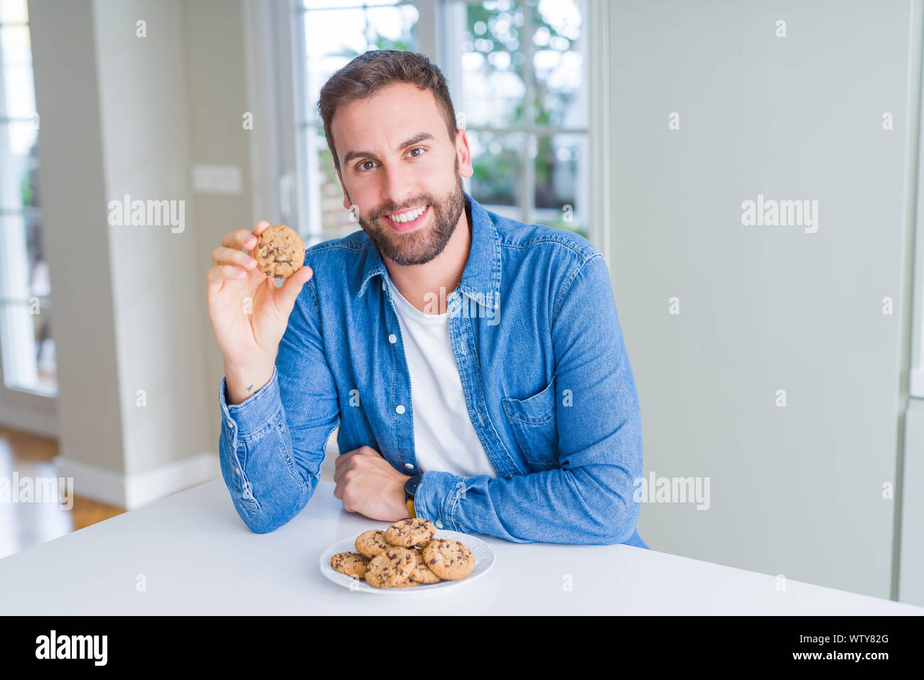 Handsome man eating chocolate chips cookies with a happy face standing ...