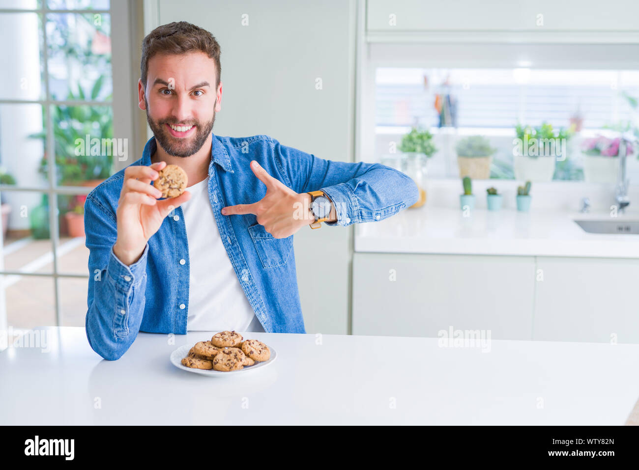Handsome man eating chocolate chips cookies with surprise face pointing ...