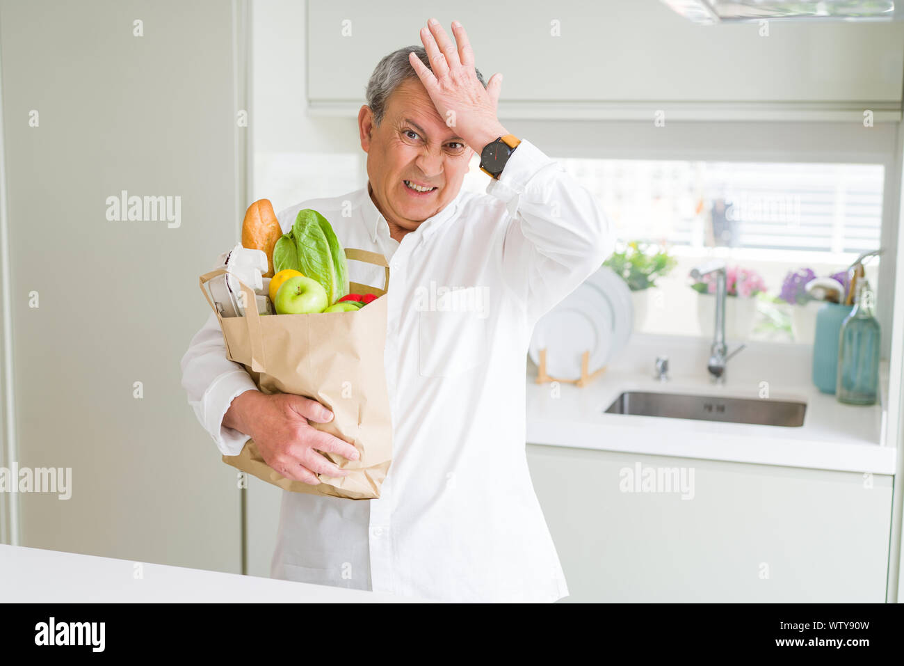 Handsome senior man holding a paper bag of fresh groceries at the kitchen stressed with hand on ...