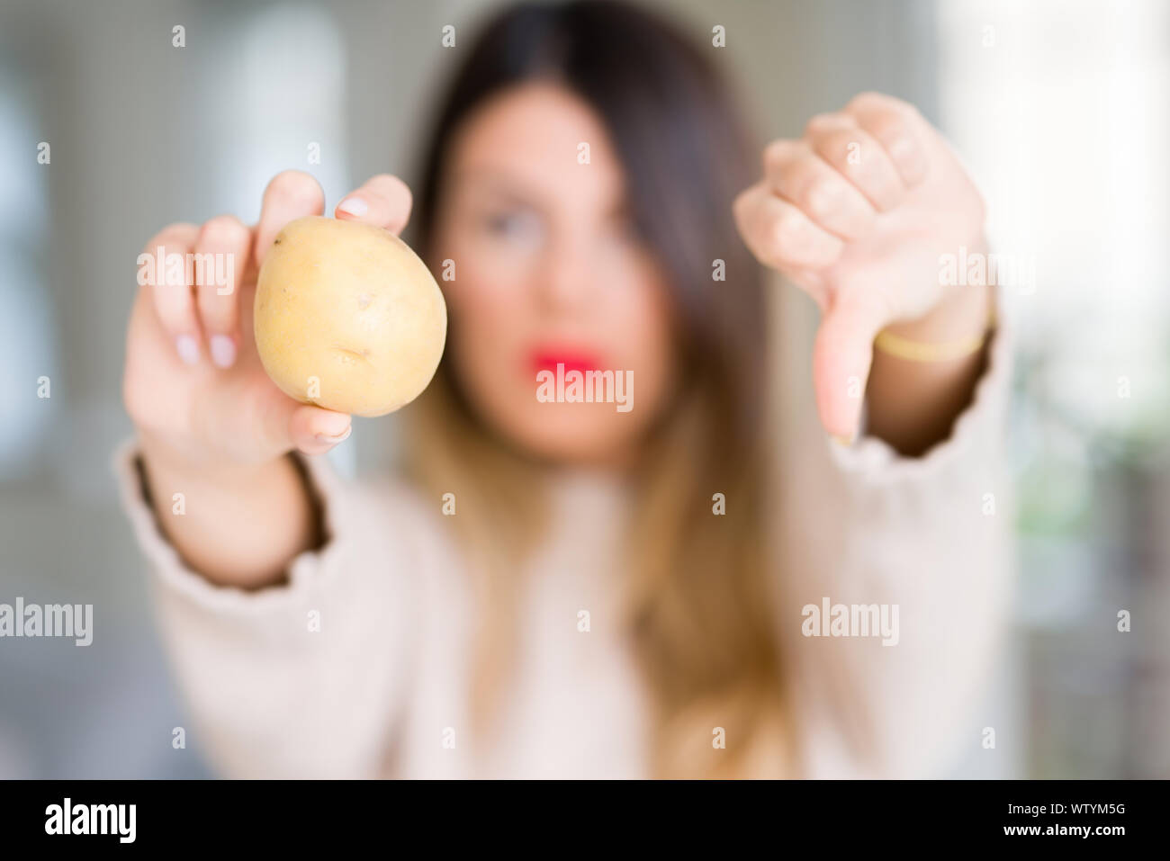 Young beautiful woman holding fresh potato at home with angry face ...