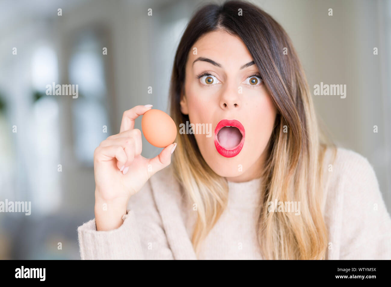 Young beautiful woman holding fresh egg at home scared in shock with a surprise face, afraid and ...