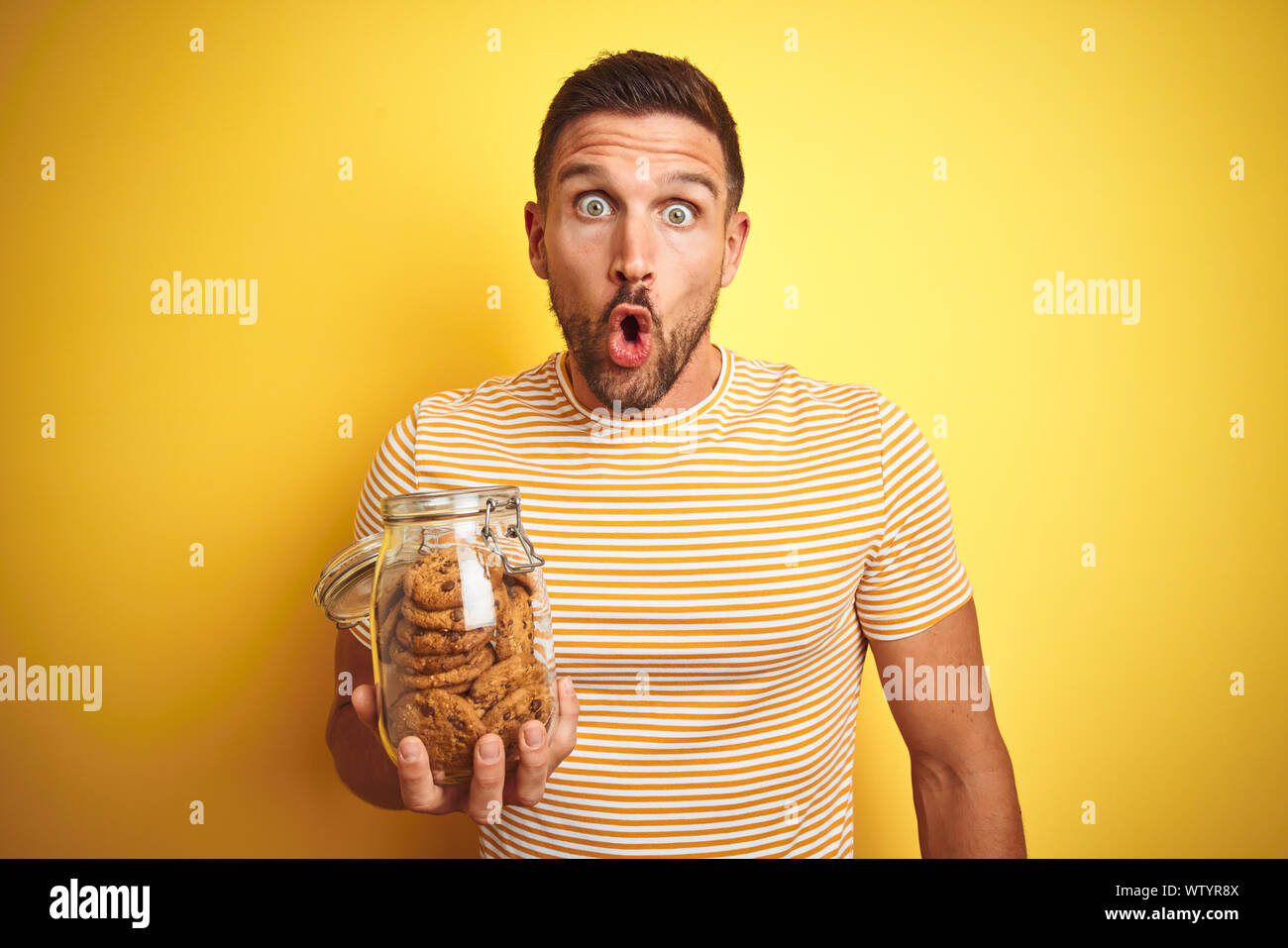 Young handsome man holding a jar of cookies over yellow isolated ...