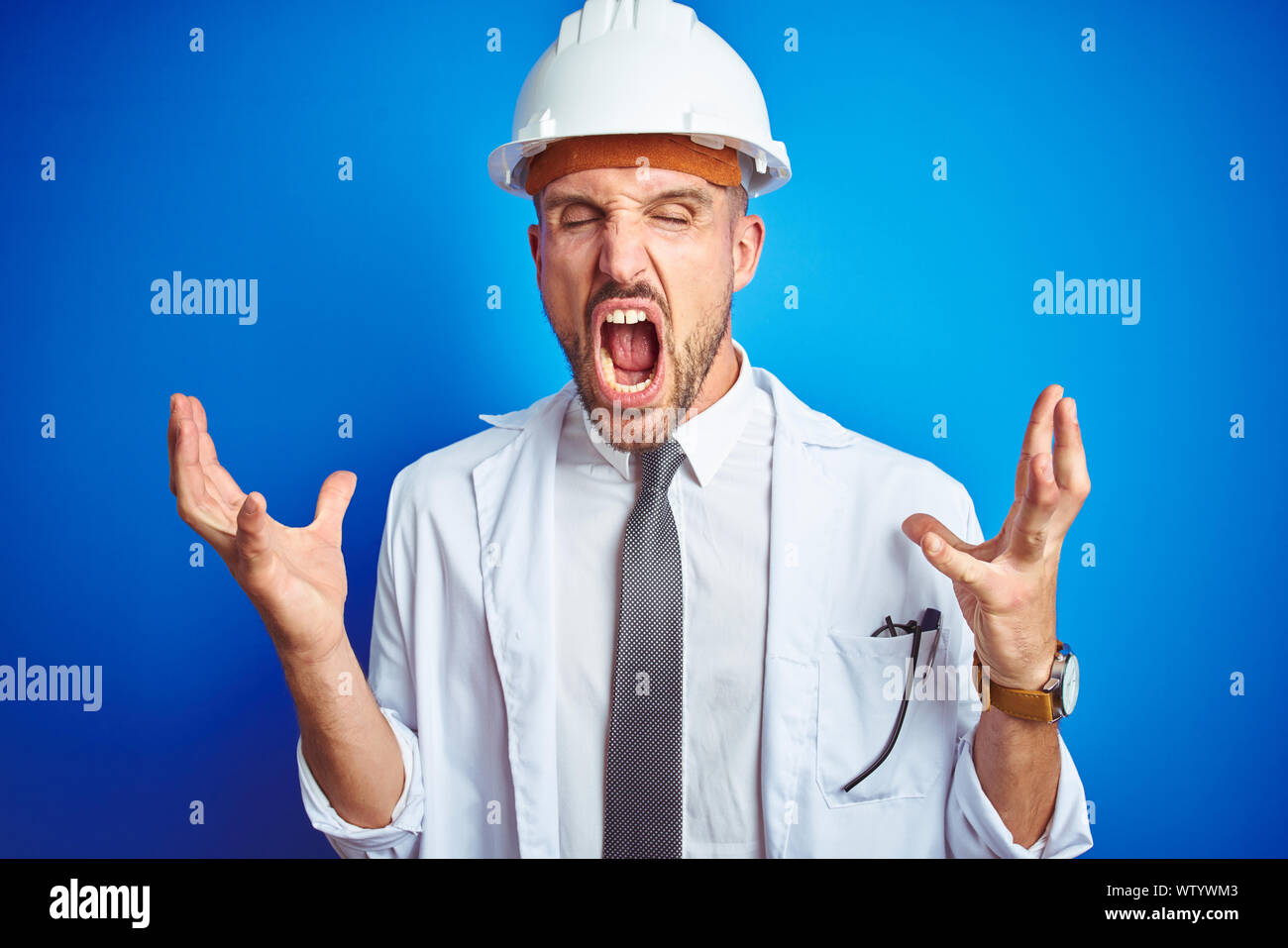 Young handsome engineer man wearing safety helmet over blue isolated ...