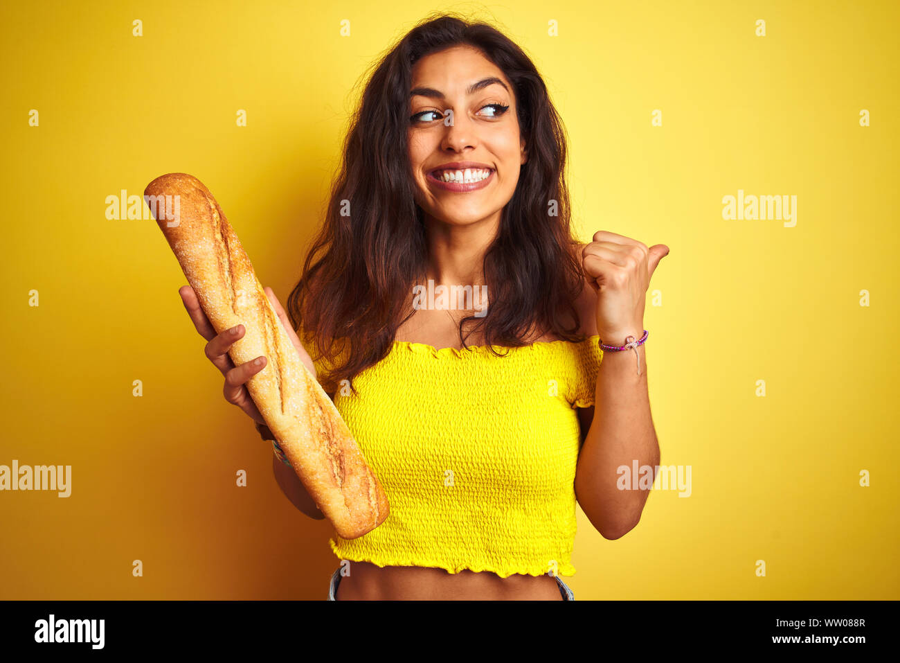 Young beautiful baker woman holding bread standing over isolated yellow ...