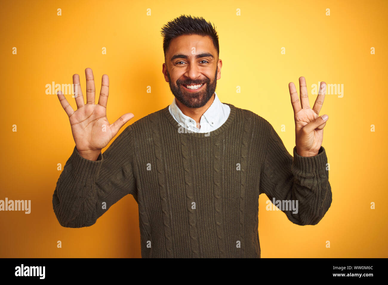 Young indian man wearing green sweater and shirt standing over isolated yellow background ...