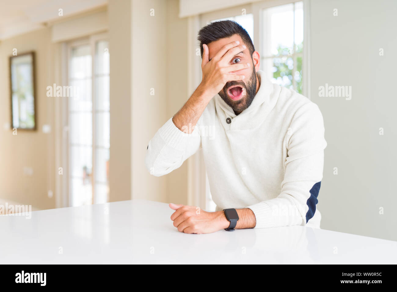 Handsome hispanic man wearing casual white sweater at home peeking in ...