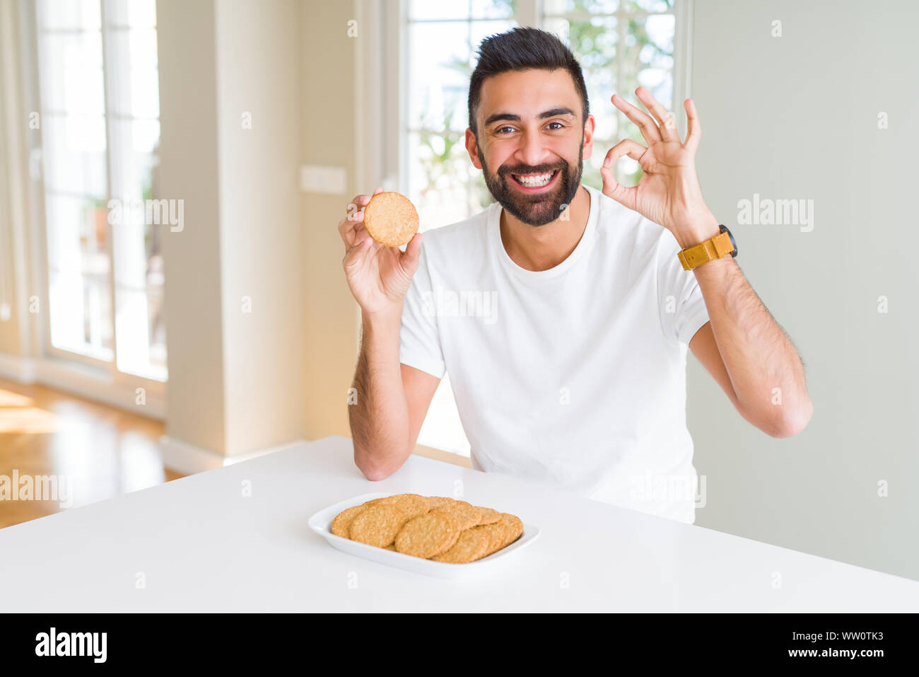 Handsome hispanic man eating healthy whole grain biscuits doing ok sign ...