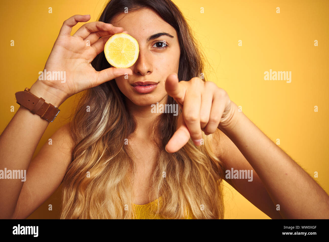 Young beautiful woman holding a lemon on eye over yellow isolated ...