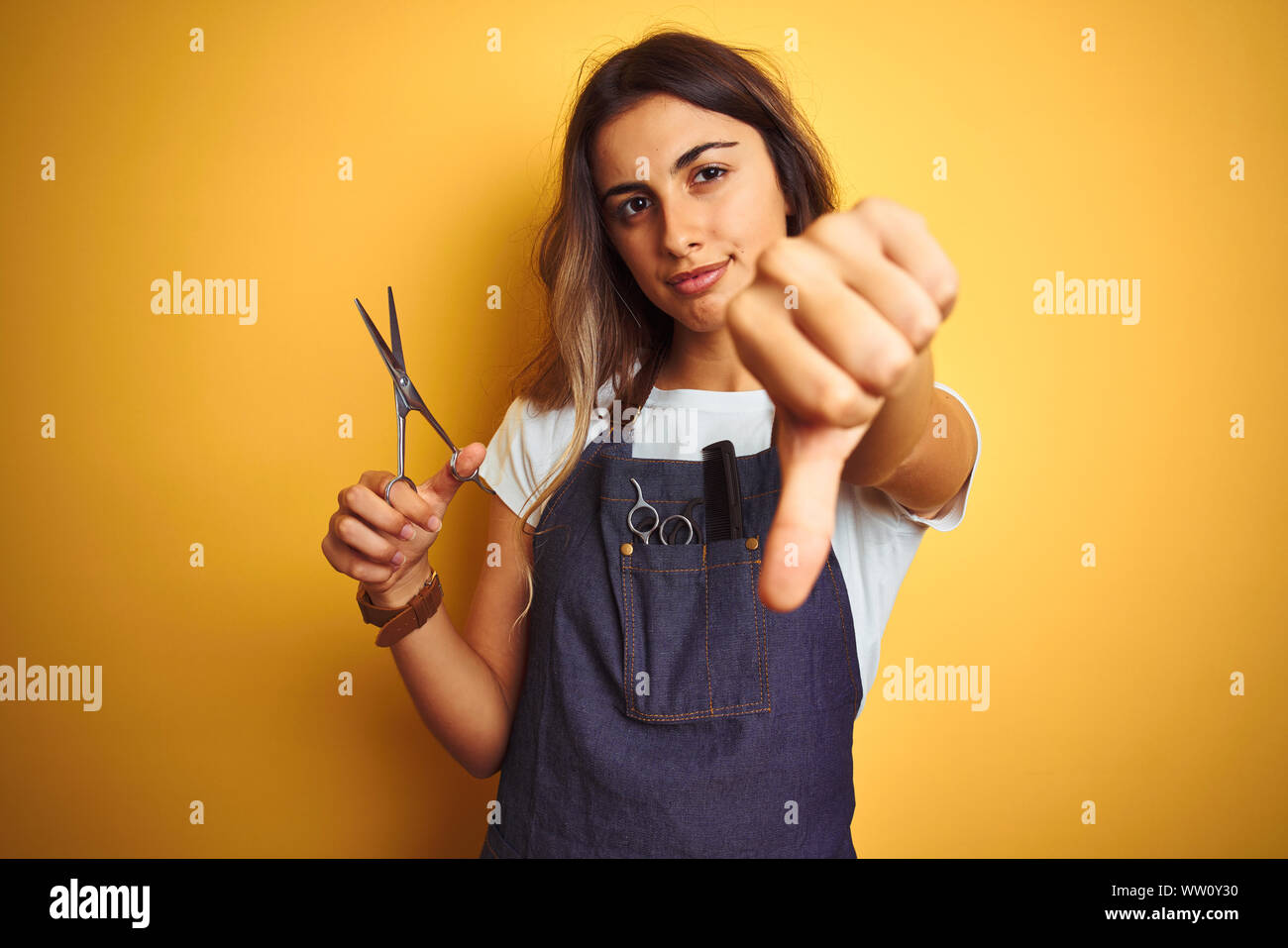 Young beautiful hairdresser woman holding scissors over yellow isolated background with angry ...