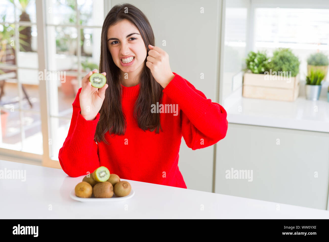 Beautiful young woman eating half fresh green kiwi annoyed and ...