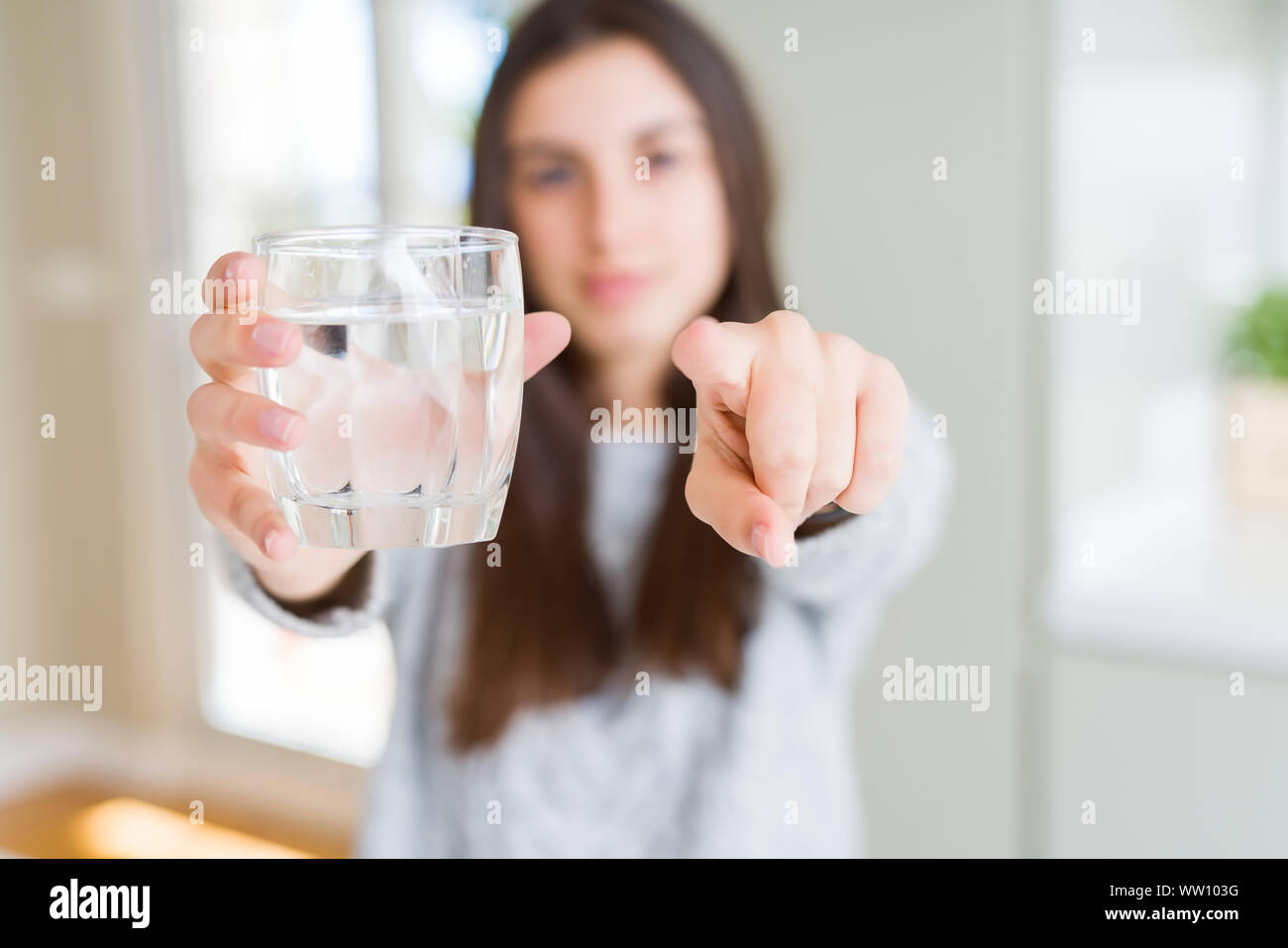 Beautiful young woman drinking a fresh glass of water pointing with finger to the camera and to ...