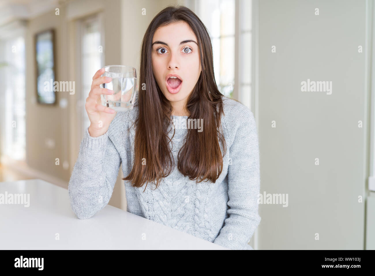 Beautiful young woman drinking a fresh glass of water scared in shock ...