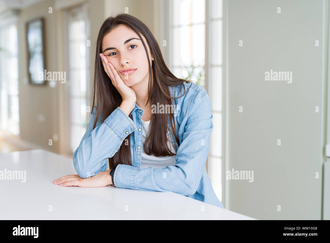 Beautiful young woman sitting on white table at home thinking looking ...