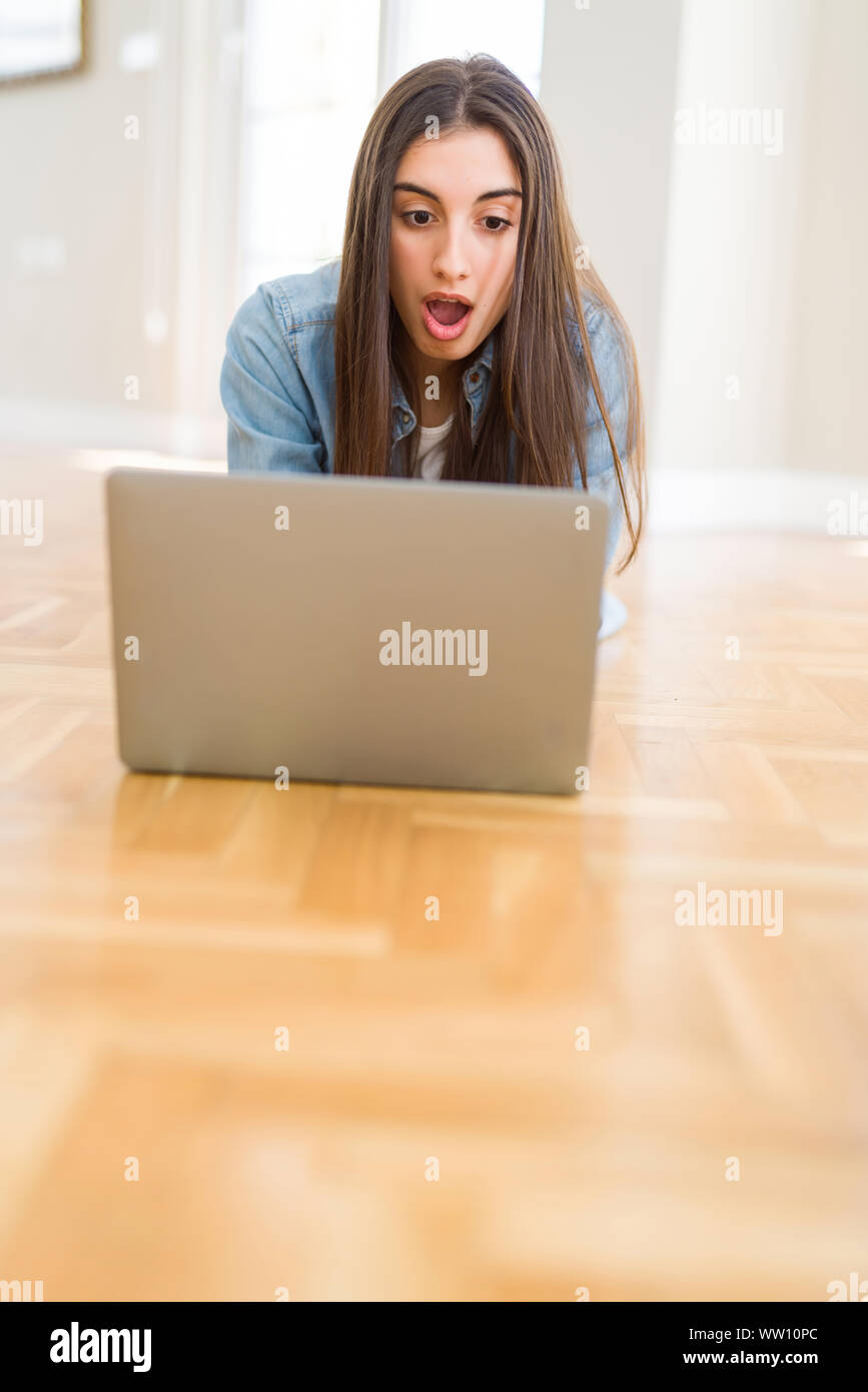 Beautiful young woman laying on the floor using laptop scared in shock ...