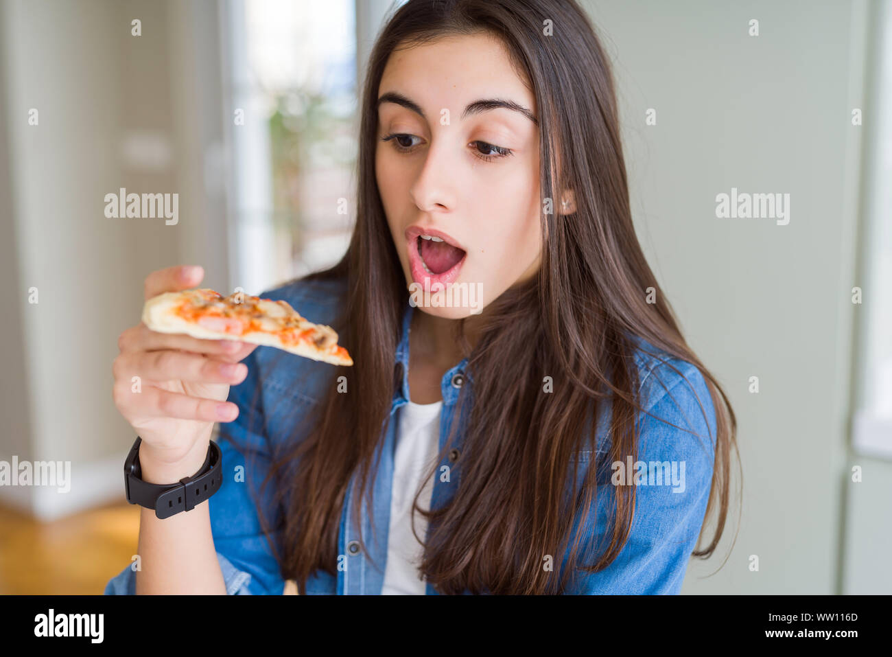 Beautiful young woman eating a slice of tasty pizza scared in shock ...