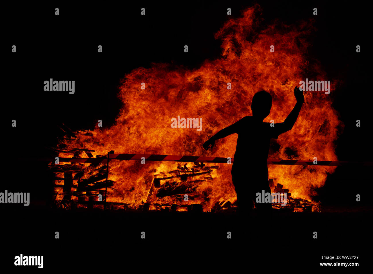 Figure with arms raised silhouetted against huge orange fire throwing ...