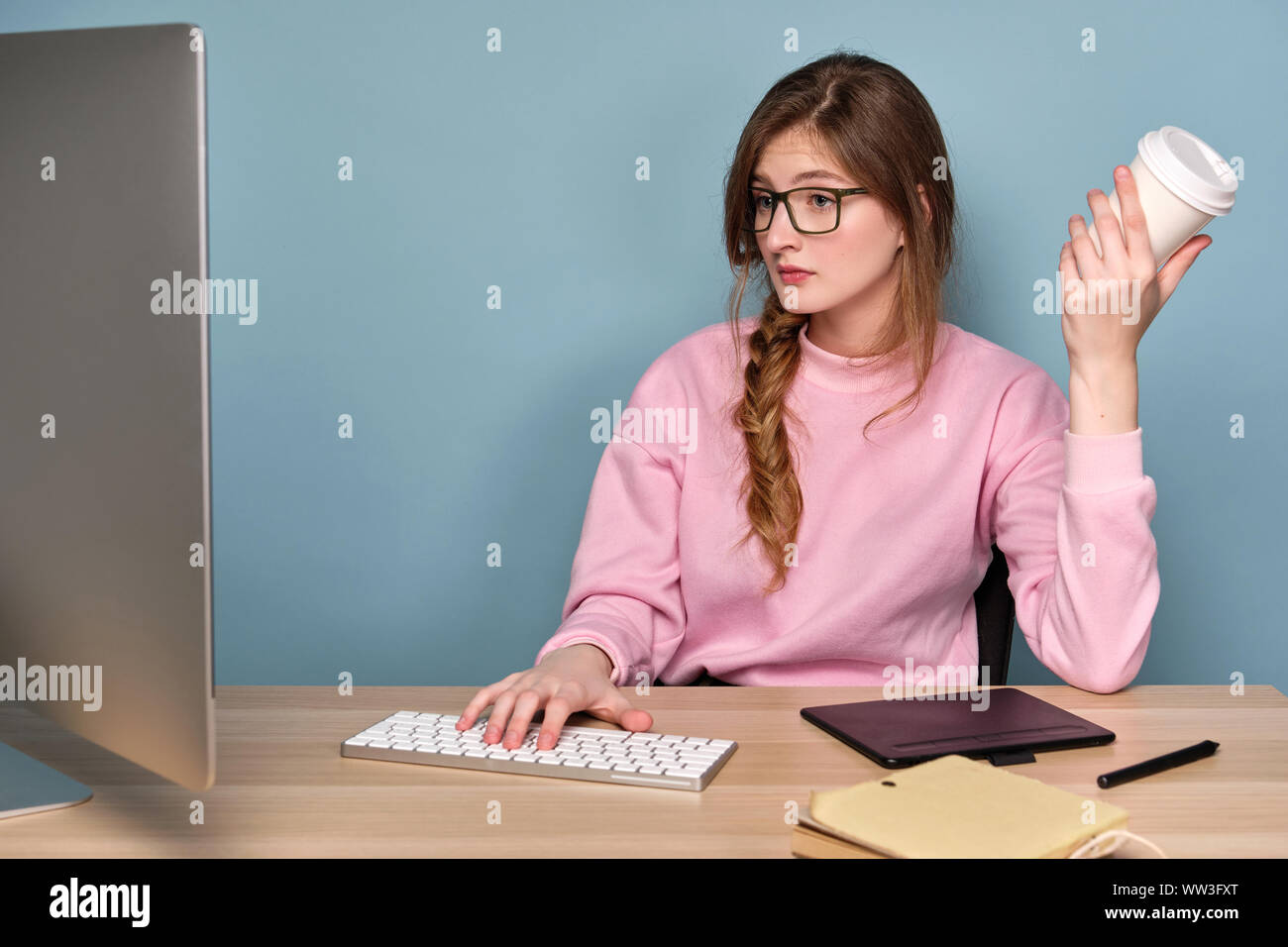 A girl in a pink sweater and glasses sits at a computer and looks in ...