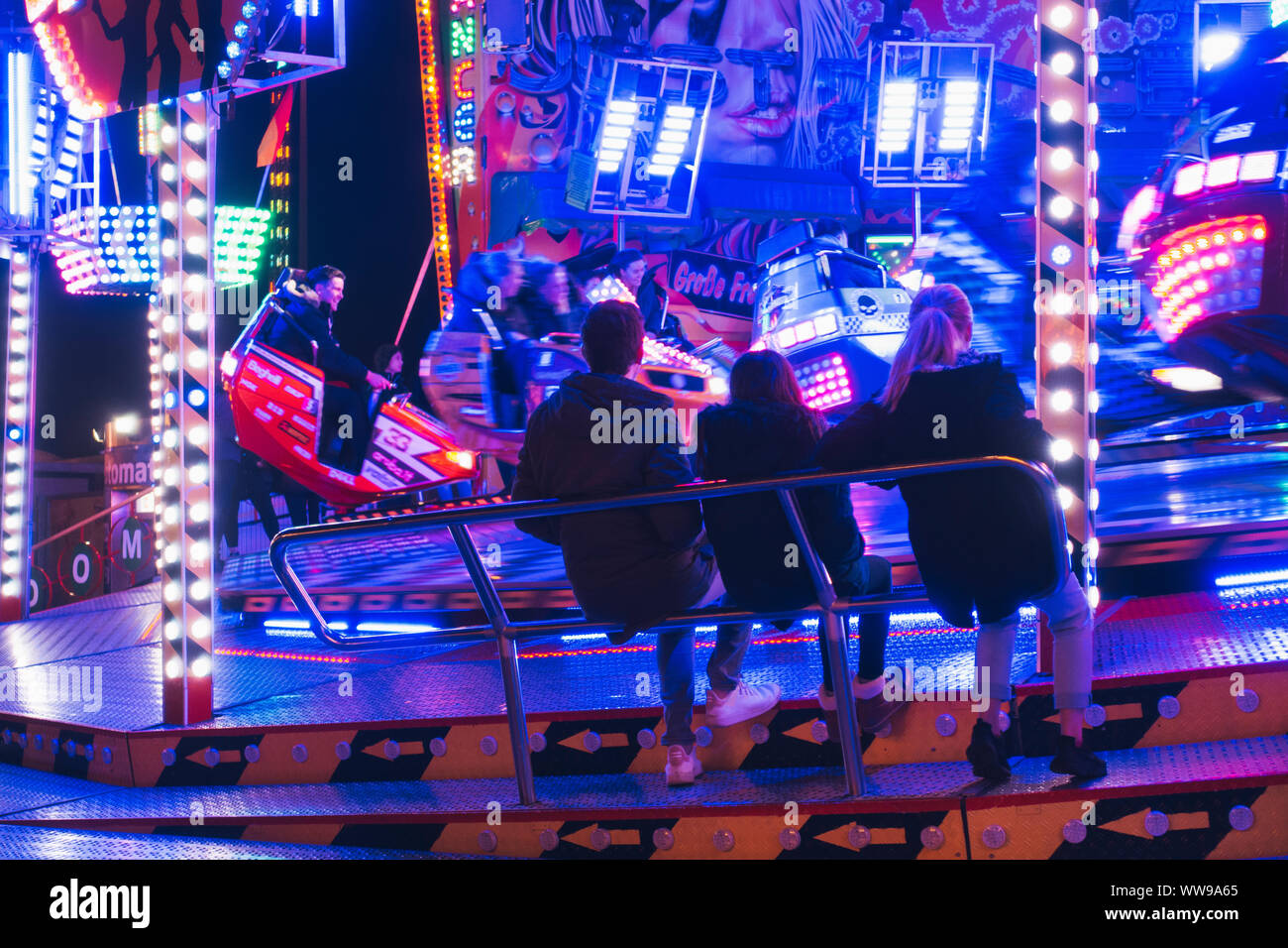 Teenagers watch the cars on a spinning fairground ride zip past them at ...