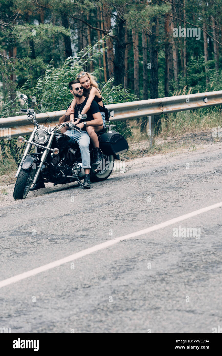young couple of bikers hugging on black motorcycle on road near green ...
