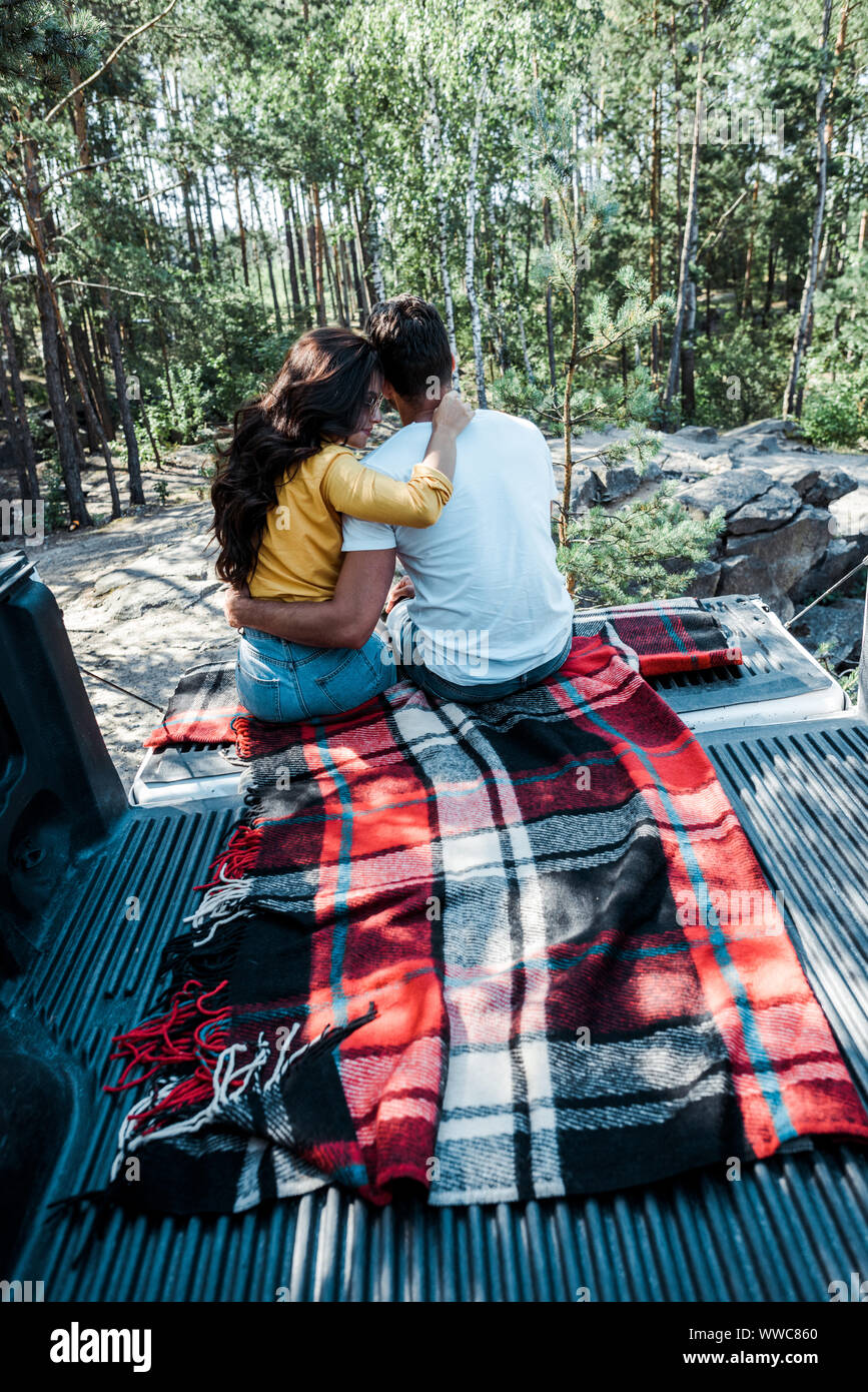 back view of woman and man hugging while sitting in car trunk in woods ...