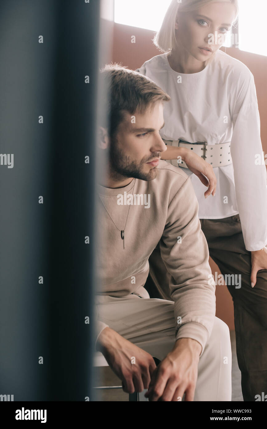 bearded man sitting on chair and attractive blonde woman near curtain ...