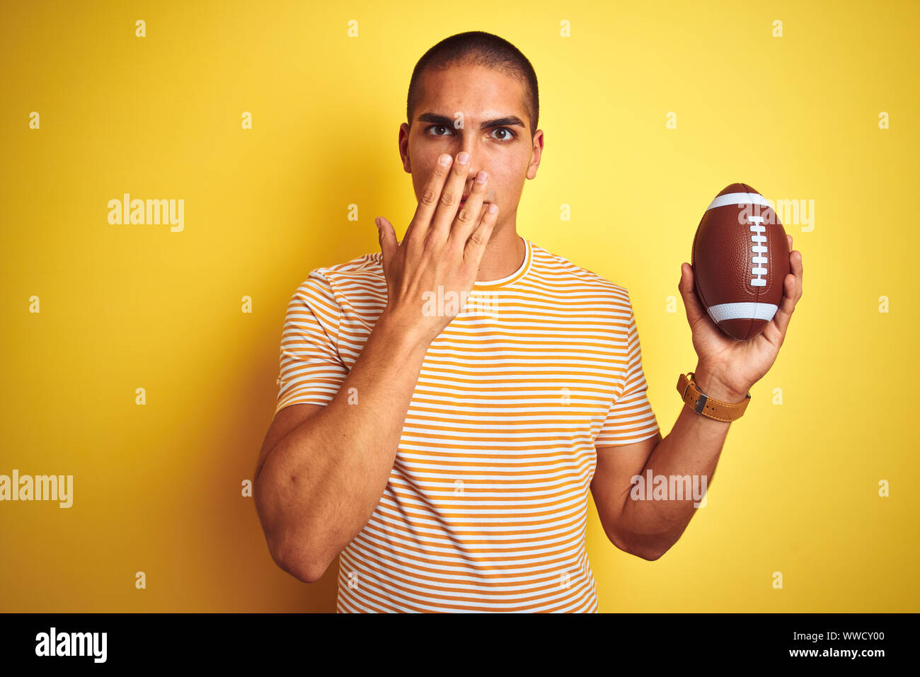 Young rugby player man holding a football ball over yellow isolated ...