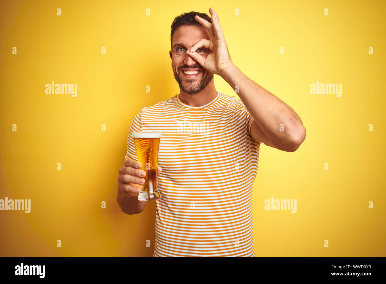Young handsome man drinking a pint glass of beer over isolated yellow ...