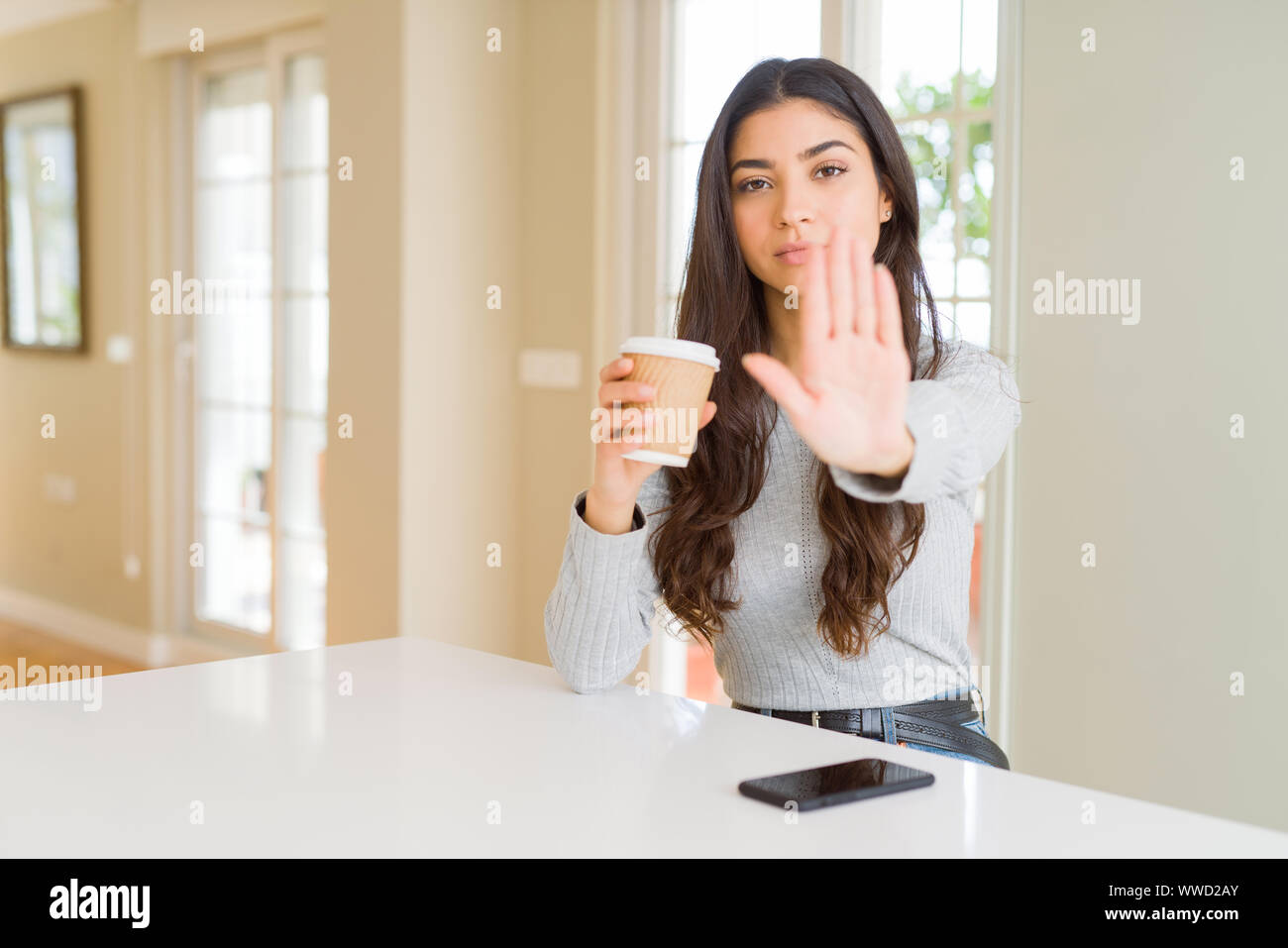 Young woman drinking a cup of coffee at home with open hand doing stop ...