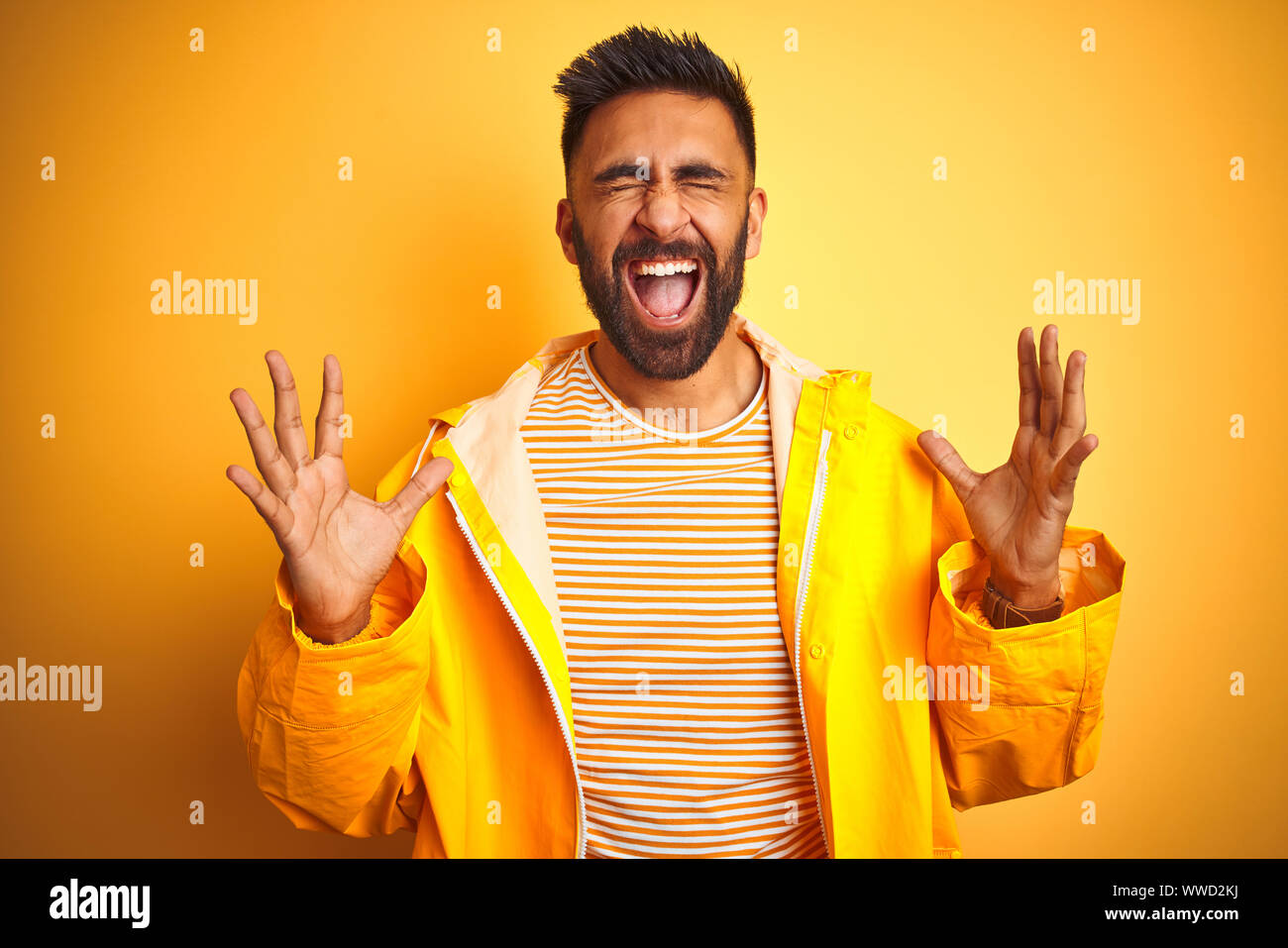 Young indian man wearing raincoat standing over isolated yellow ...