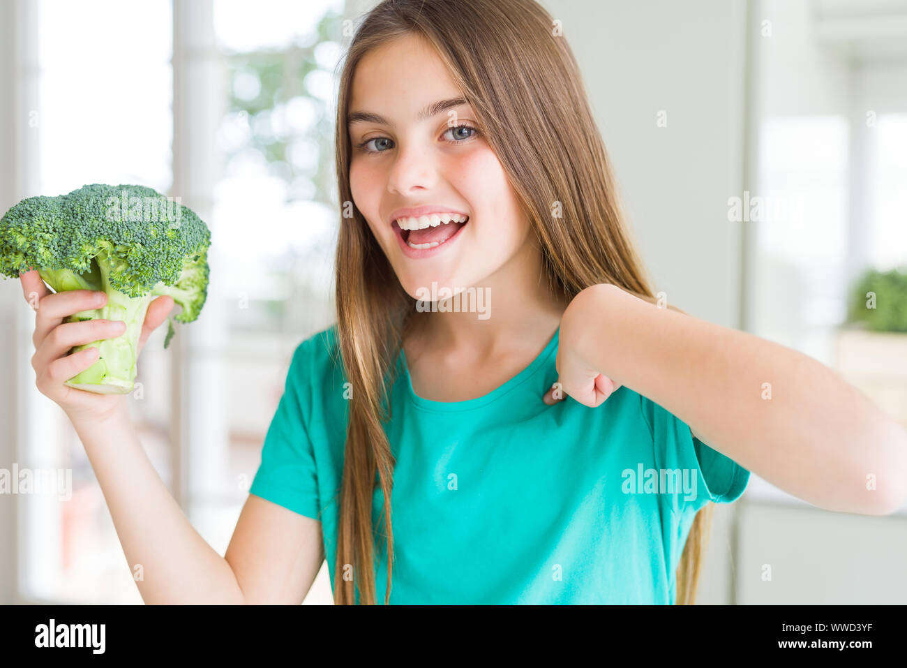 Beautiful young girl eating fresh broccoli with surprise face pointing ...