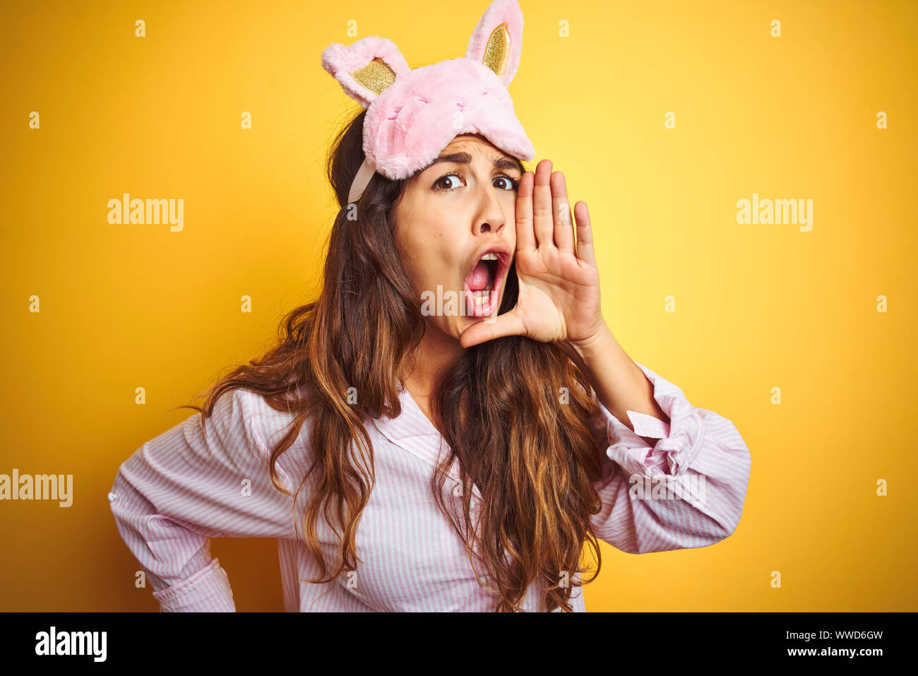 Young woman wearing pajama and sleep mask standing over yellow isolated ...