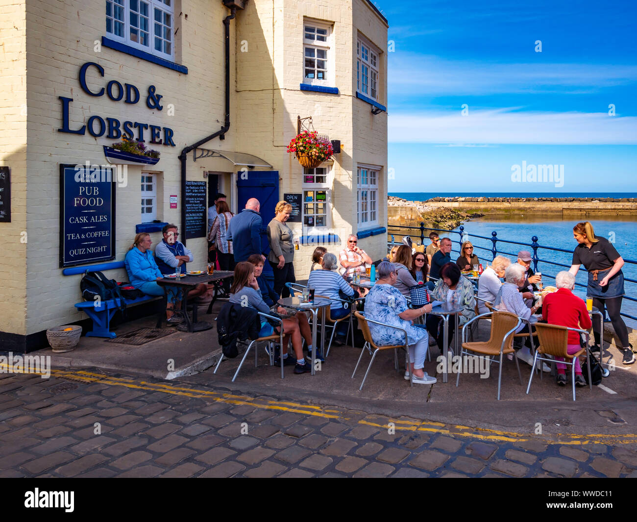 A crowd of people eating outside the Cod and Lobster pub on the harbour ...
