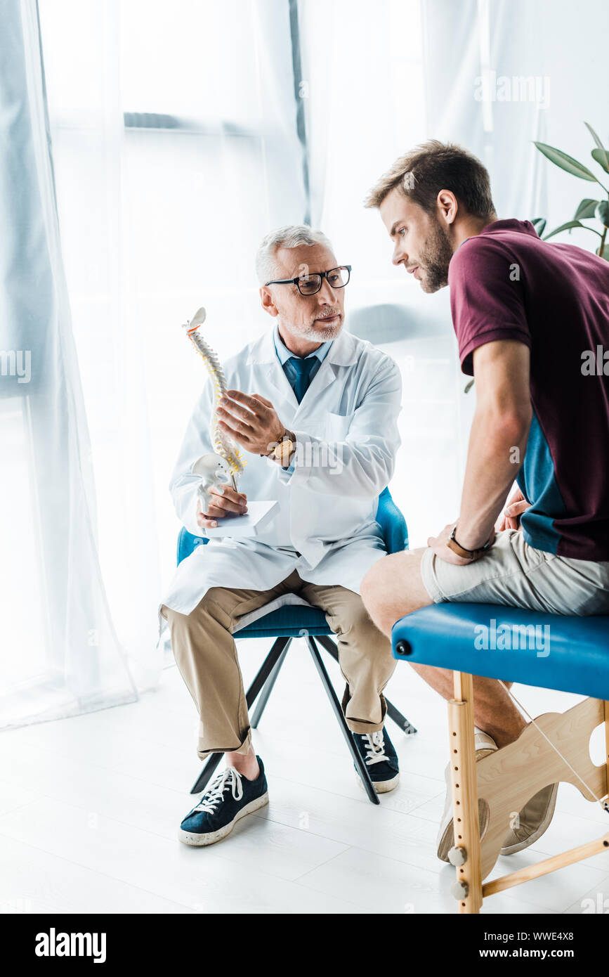 bearded doctor in glasses holding spine model and looking at patient ...