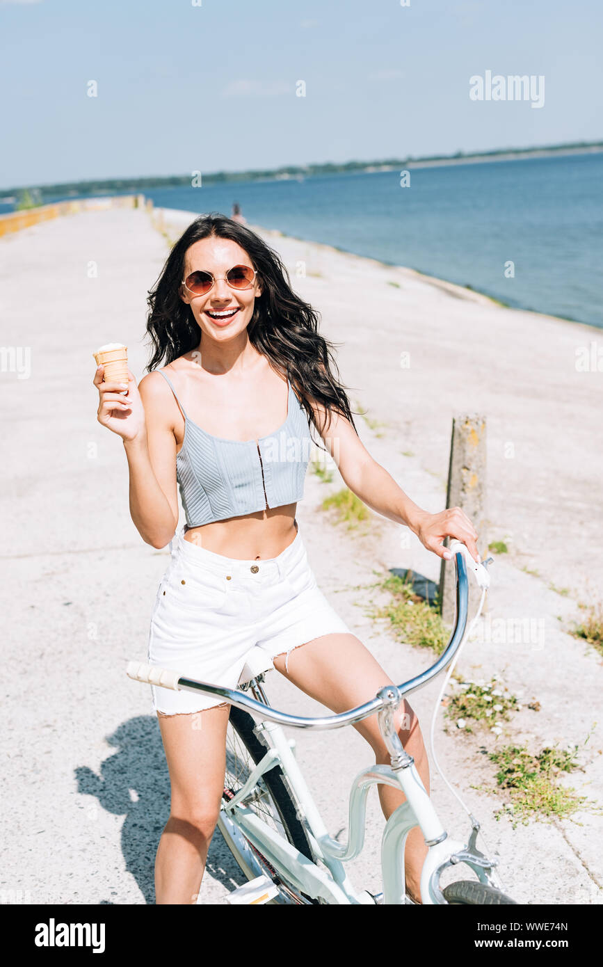 happy brunette girl with ice cream riding bike near river in summer ...