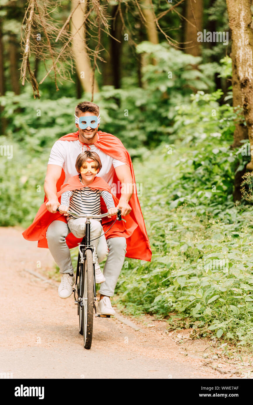 father and kid smiling while riding bicycle around forest in superhero ...