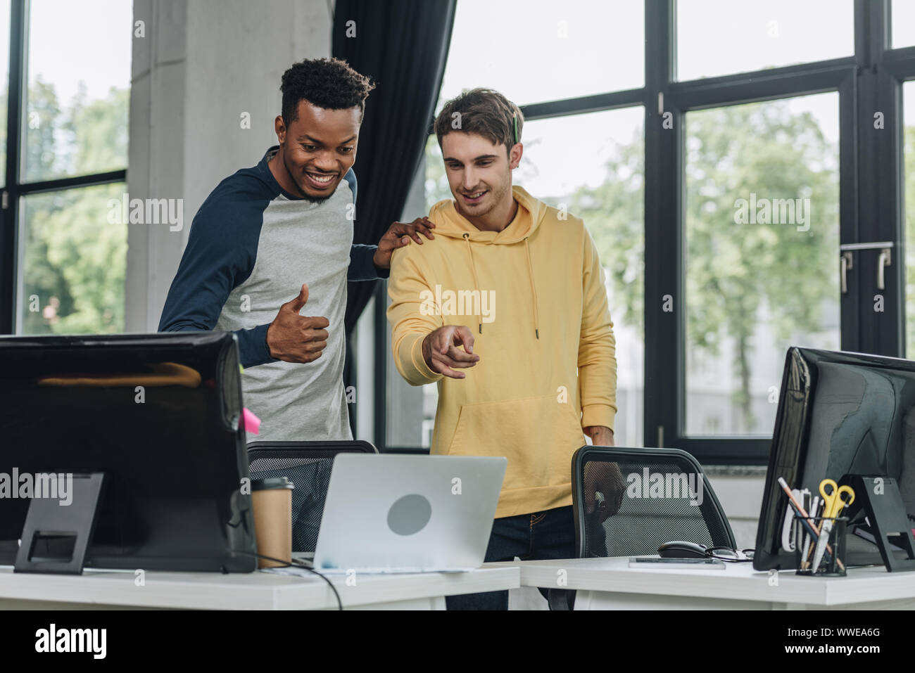 two cheerful multicultural programmers looking at laptop in office ...