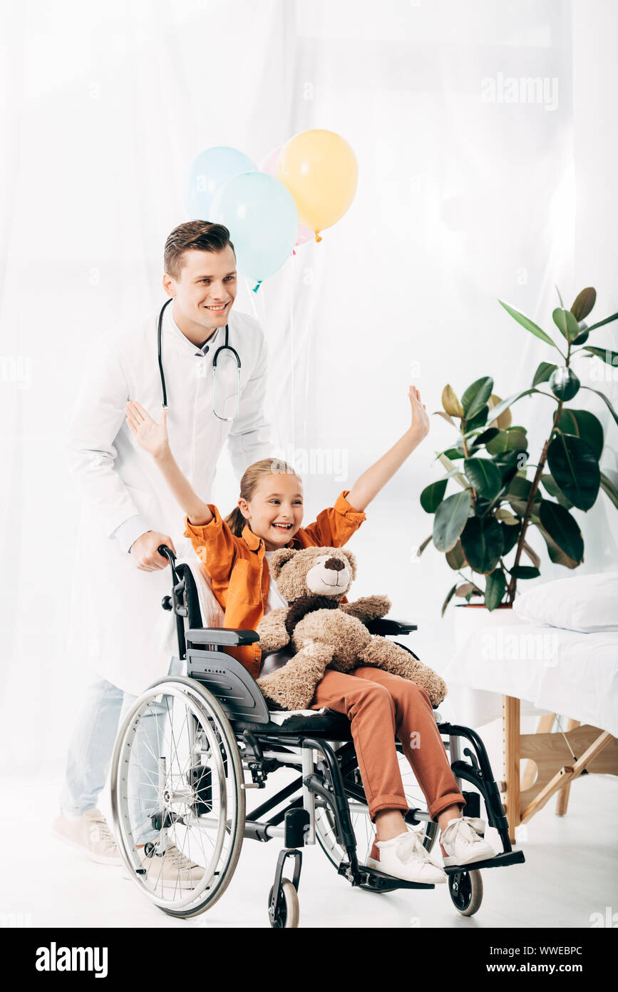 smiling pediatrist and kid with teddy bear on wheelchair in hospital ...