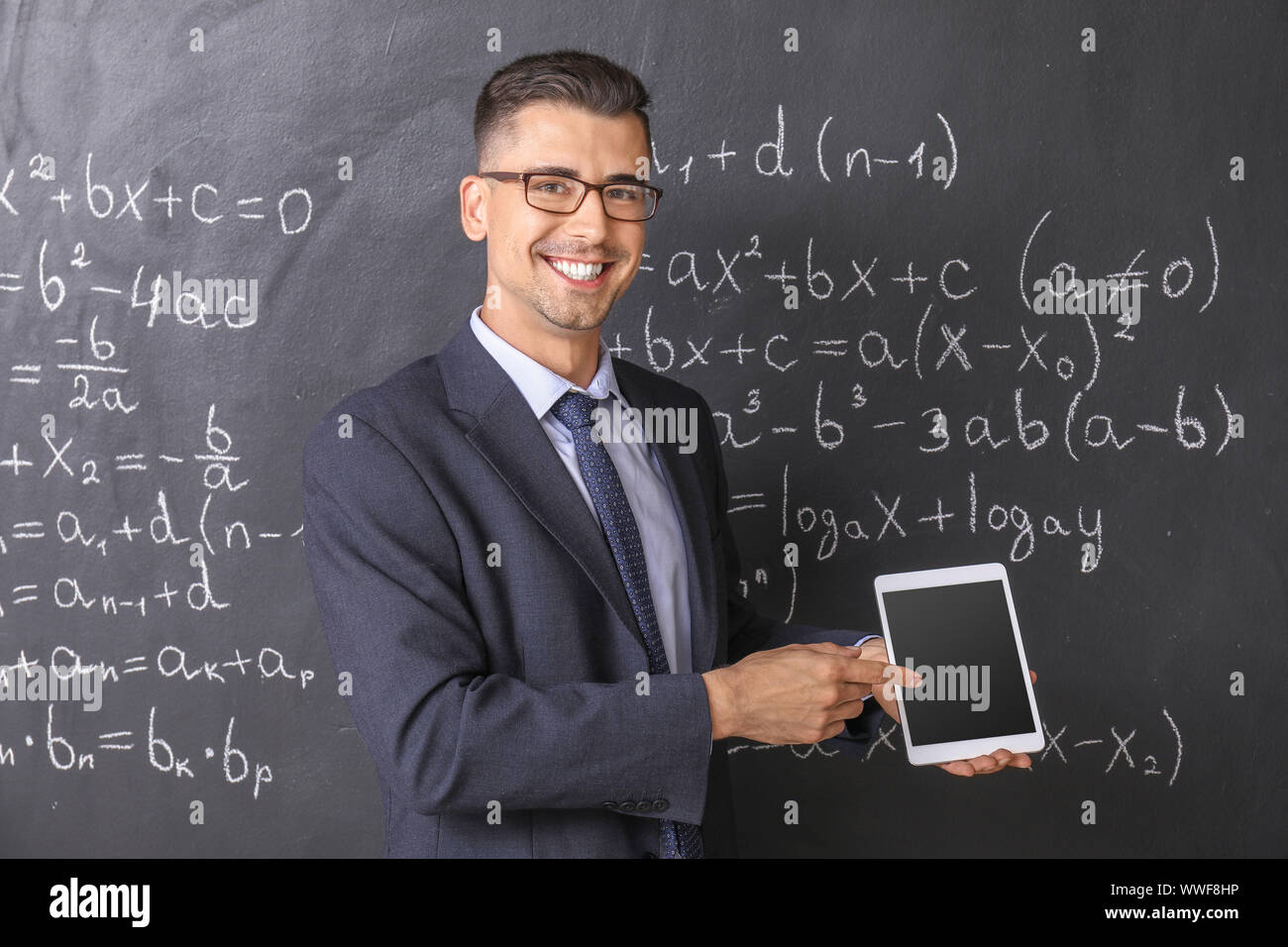 Handsome math teacher with tablet computer near blackboard in classroom ...