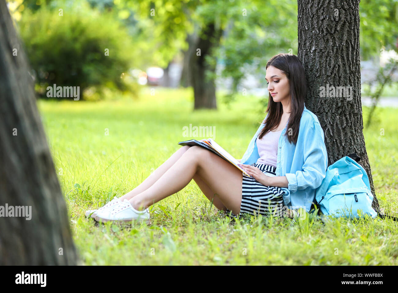 Beautiful female student sitting under tree in park Stock Photo - Alamy