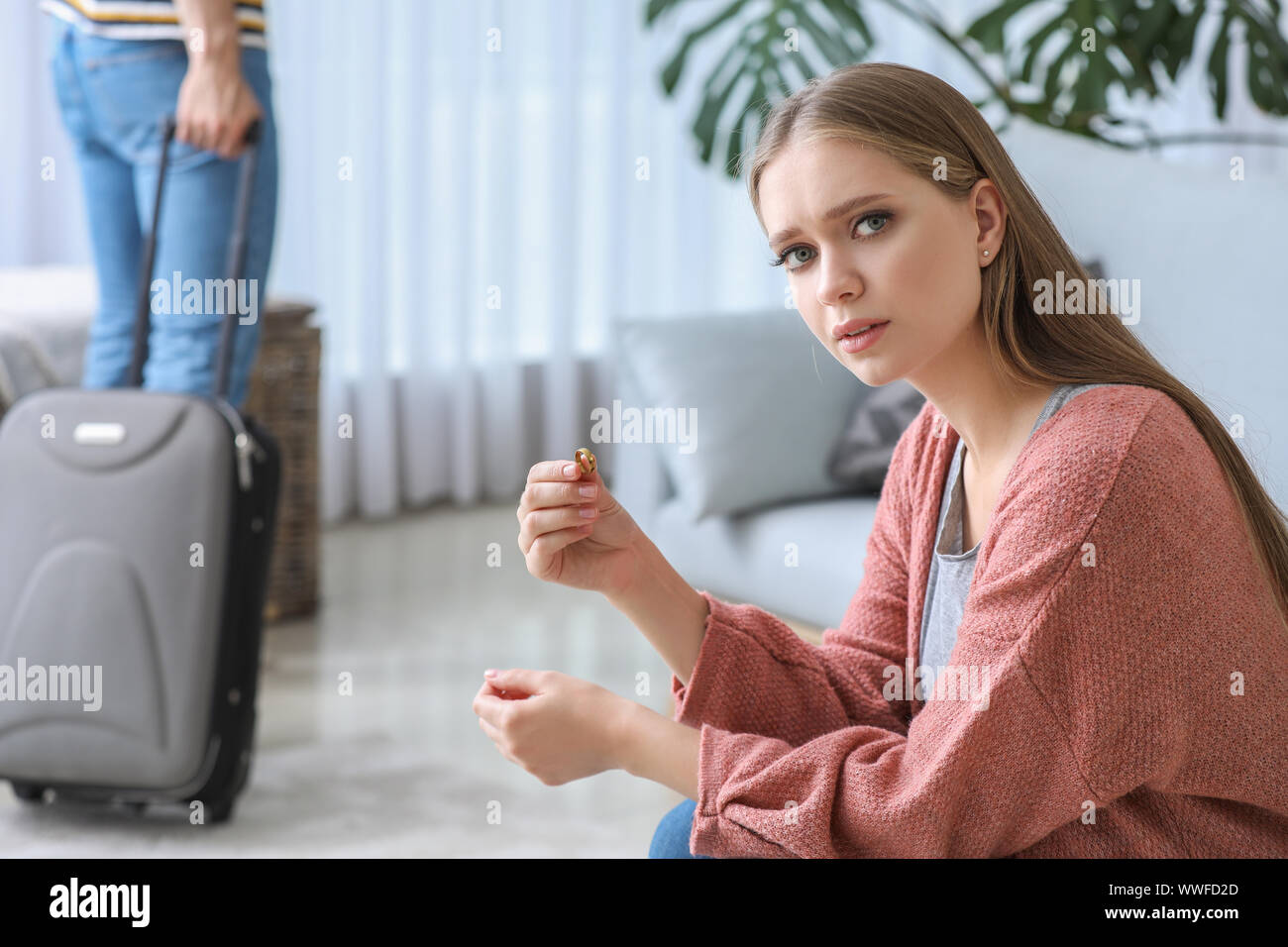 Sad young woman holding wedding ring and her husband with suitcase at ...