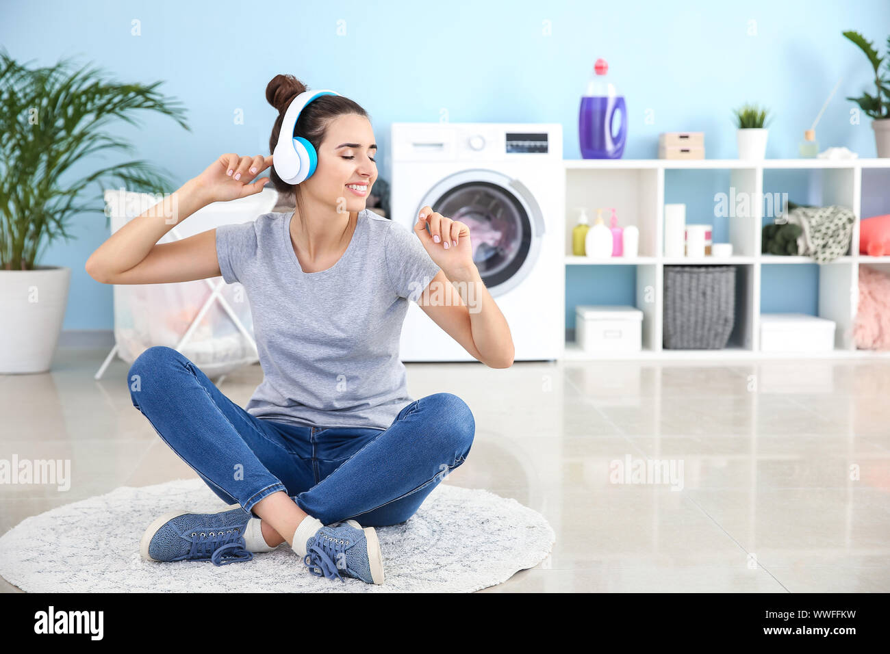 Beautiful young woman listening to music while doing laundry at home ...
