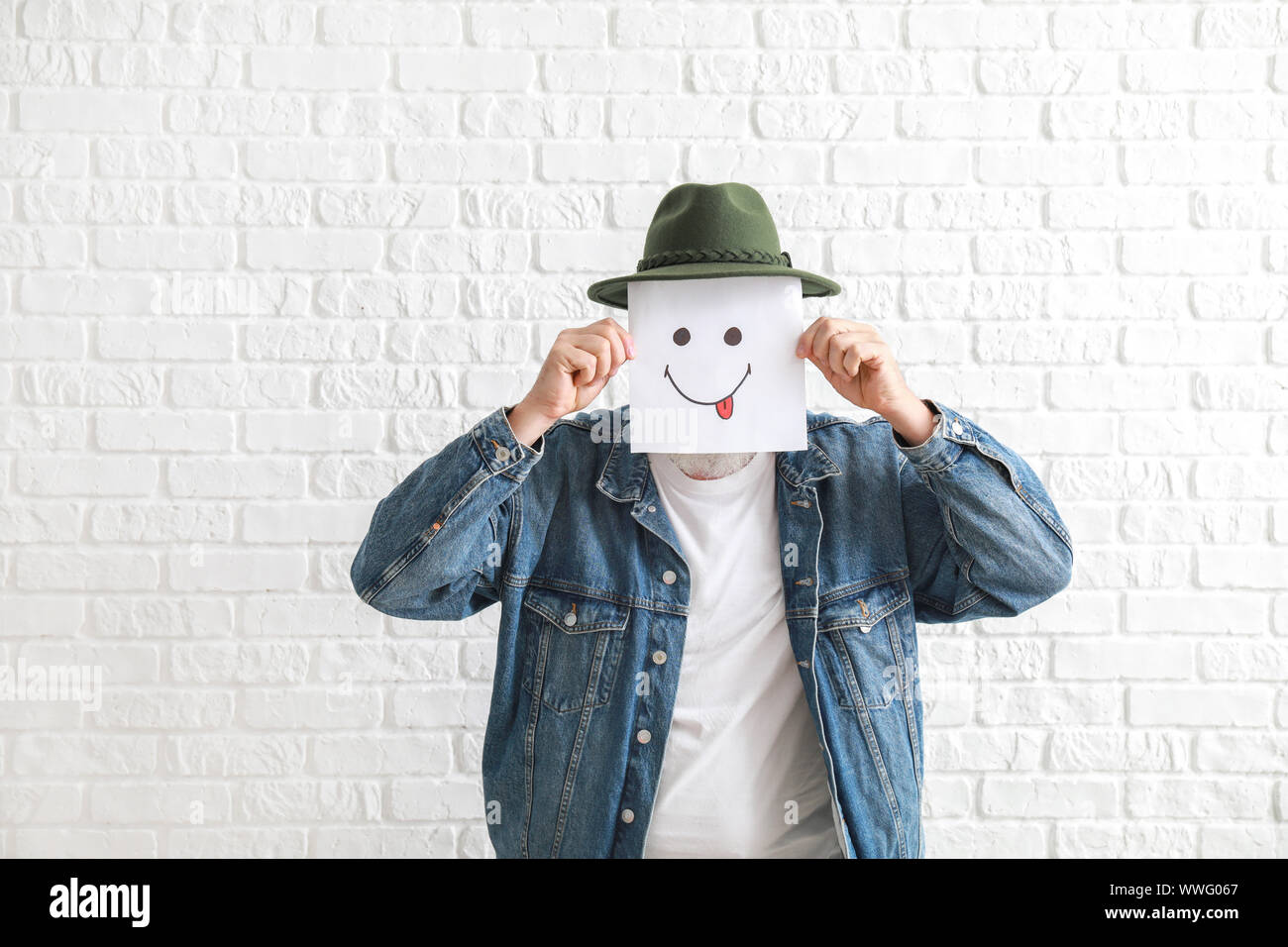 Mature man hiding face behind emoticon against white brick wall Stock ...