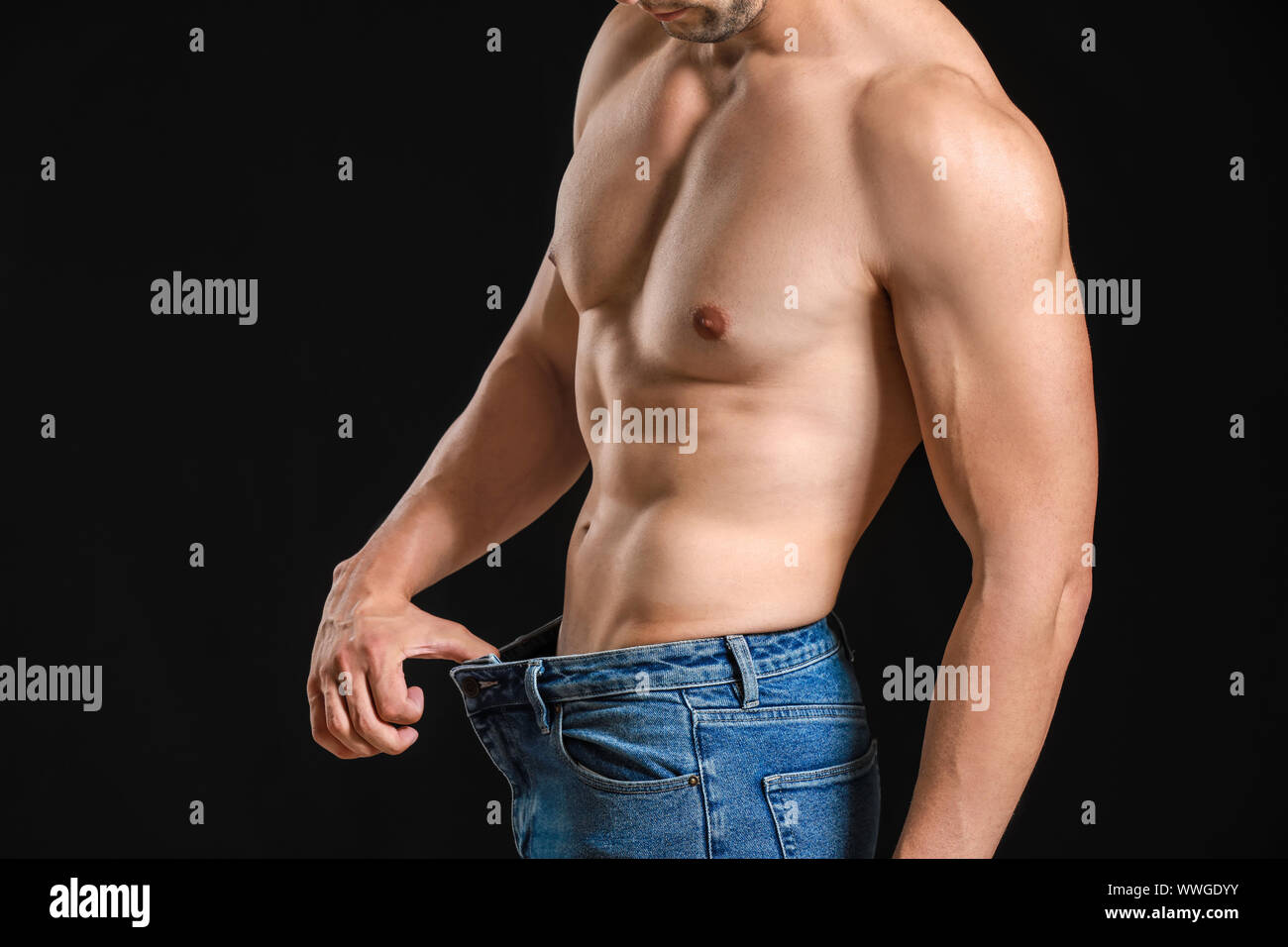 Handsome muscular man in loose jeans on dark background. Weight loss ...