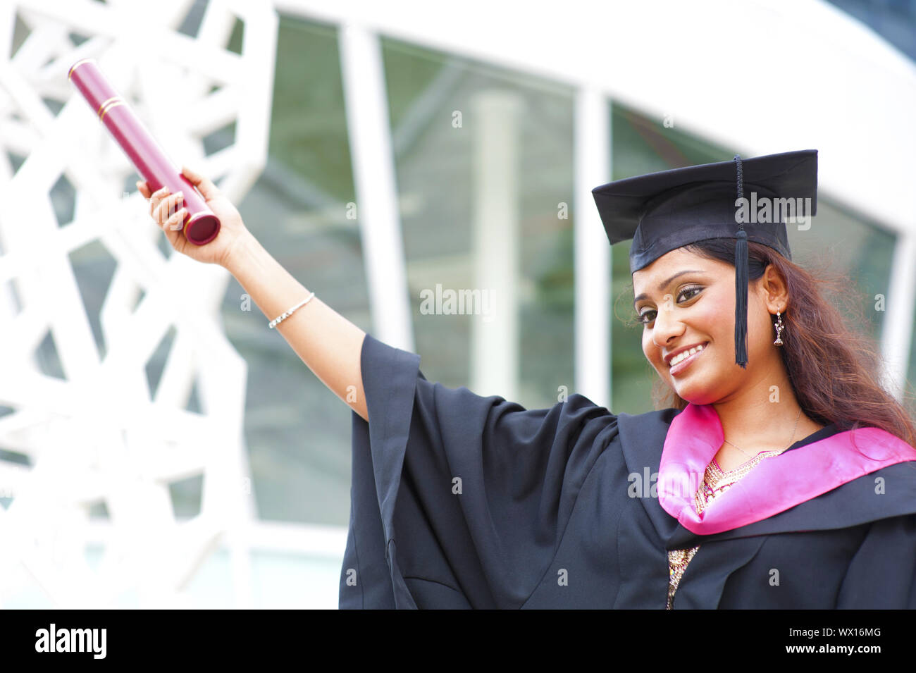 Happy young Indian in a graduation gown and cap holding her diploma ...
